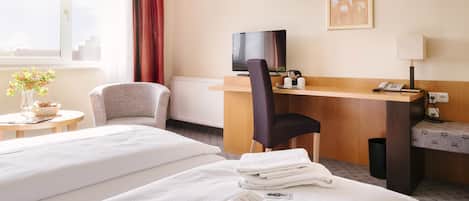 Quarto de hotel com duas camas, uma escrivaninha com cadeira, televisão, abajur e janela com cortinas.