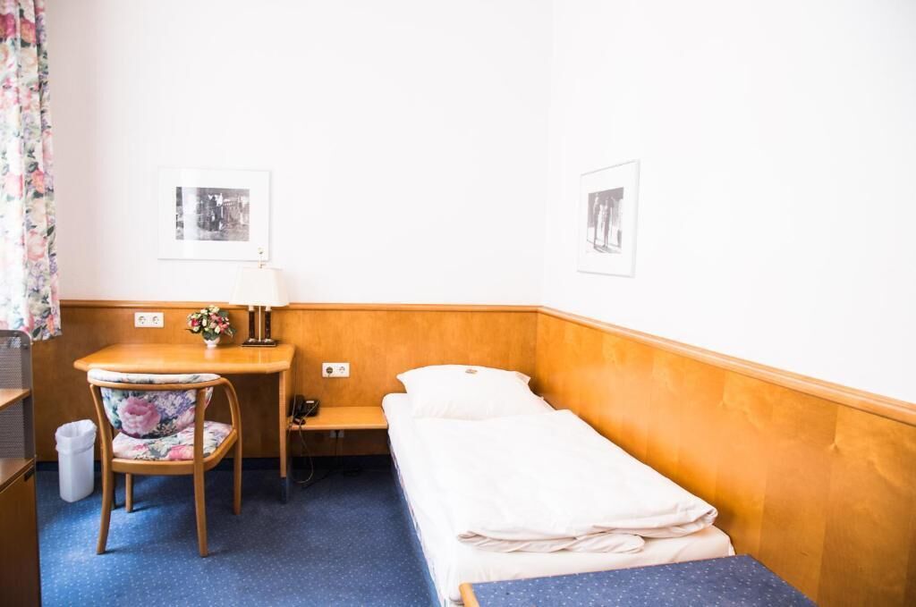 Quarto pequeno com uma cama de solteiro, cabeceira de madeira, escrivaninha com cadeira e duas imagens emolduradas na parede.