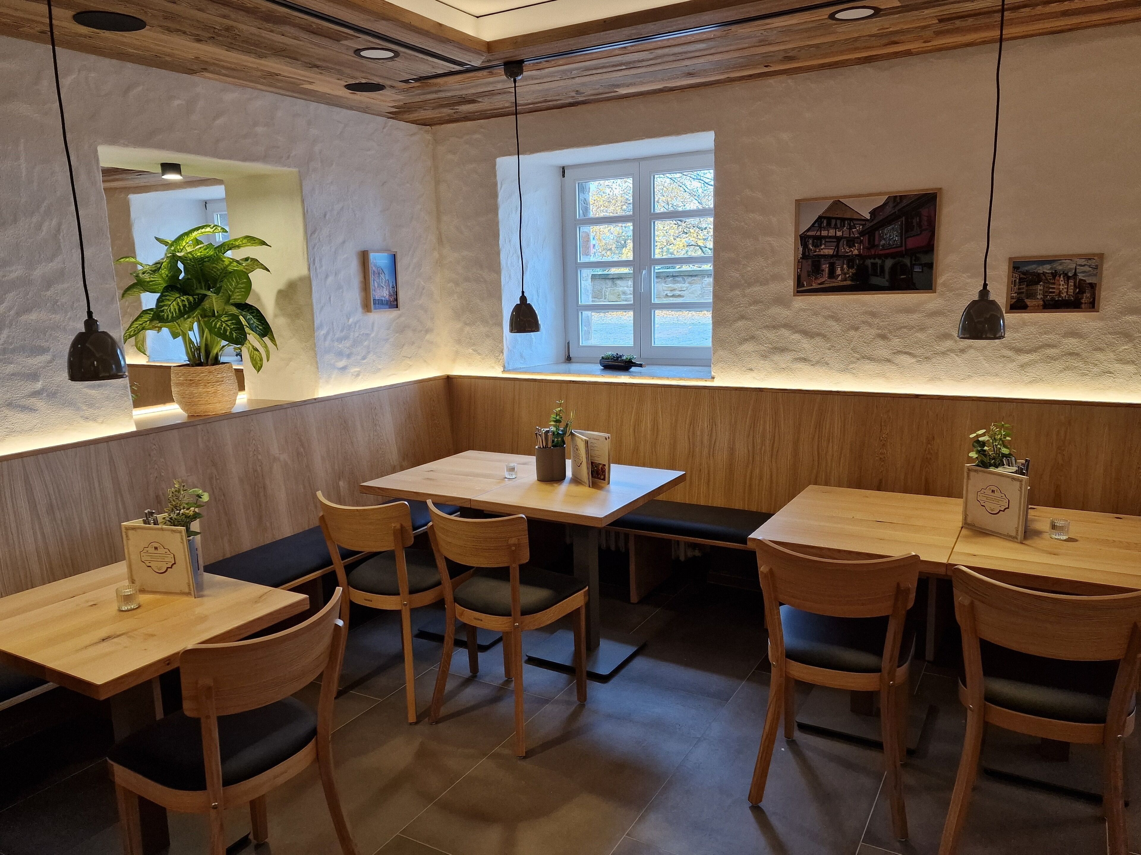 Um interior de restaurante acolhedor com mesas e cadeiras de madeira, uma planta em vaso e obras de arte nas paredes.