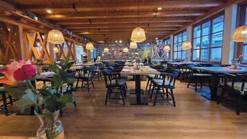 Restaurante com vigas de madeira, janelas amplas e luminárias pendentes. Mesas montadas com cadeiras e centros de mesa.