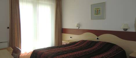 Quarto de hotel com uma cama grande, uma mesa de cabeceira, uma luminária, uma janela com cortinas e um quadro na parede.