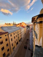 Vista da rua de uma cidade, com edifícios, céu claro e o topo de uma igreja ao longe.