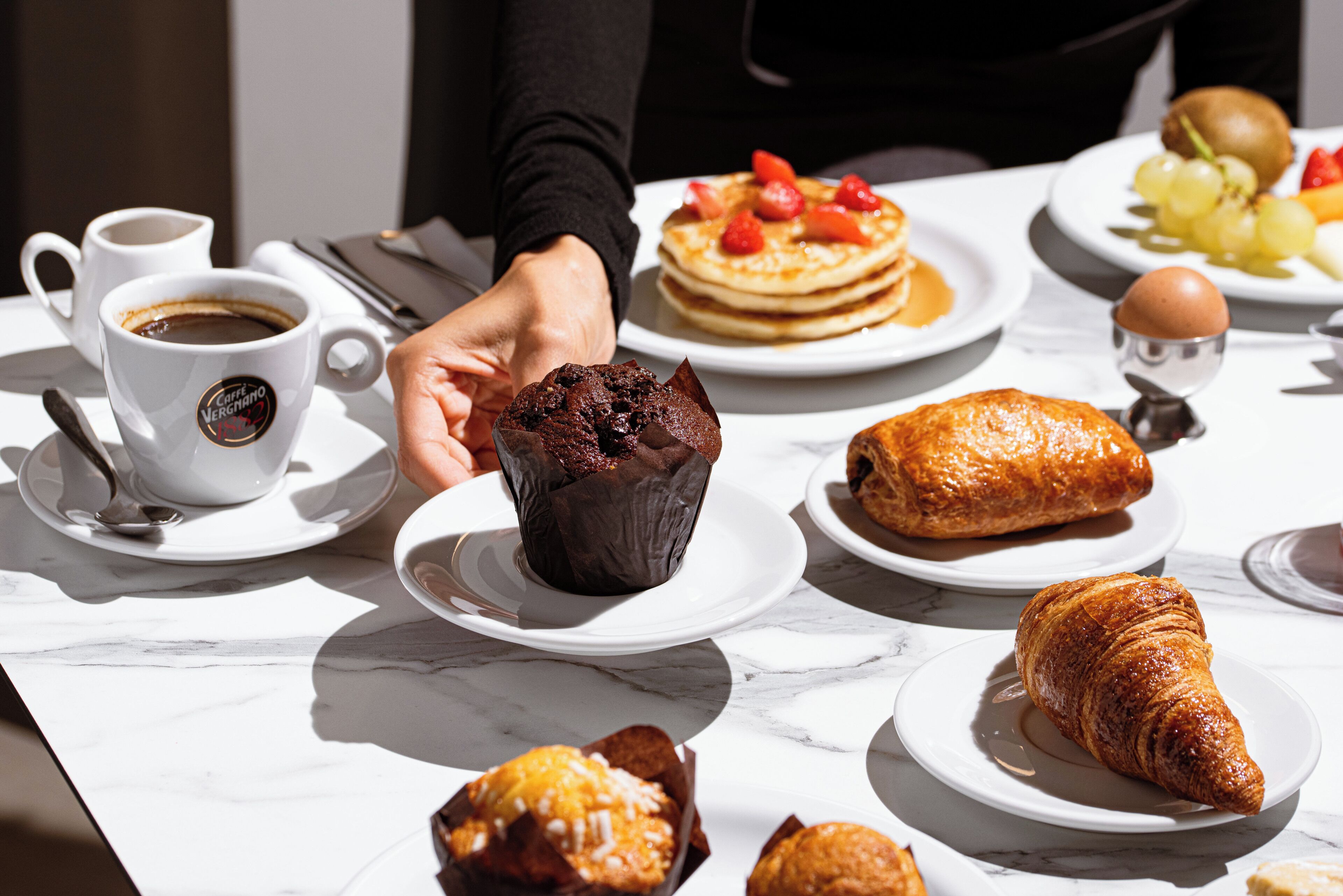 Mesa de café da manhã com diversos pães, uma xícara de café e um prato de panquecas.