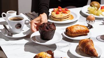 Mesa de café da manhã com diversos pães, uma xícara de café e um prato de panquecas.