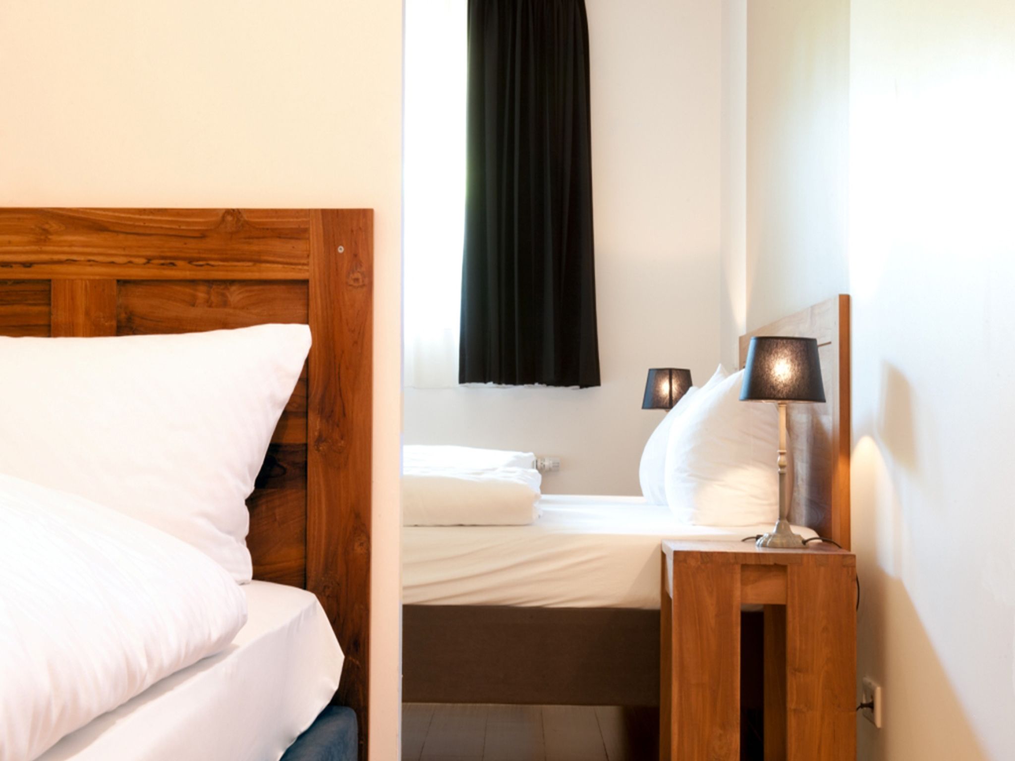 Quarto de hotel com cabeceira de madeira, cama com roupa de cama branca, mesa de cabeceira com abajur e janela com cortina preta.