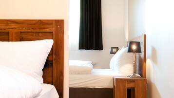 Quarto de hotel com cabeceira de madeira, cama com roupa de cama branca, mesa de cabeceira com abajur e janela com cortina preta.