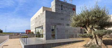 Um edifício moderno com fachada de vidro, guarda-corpo metálico e uma árvore em frente.