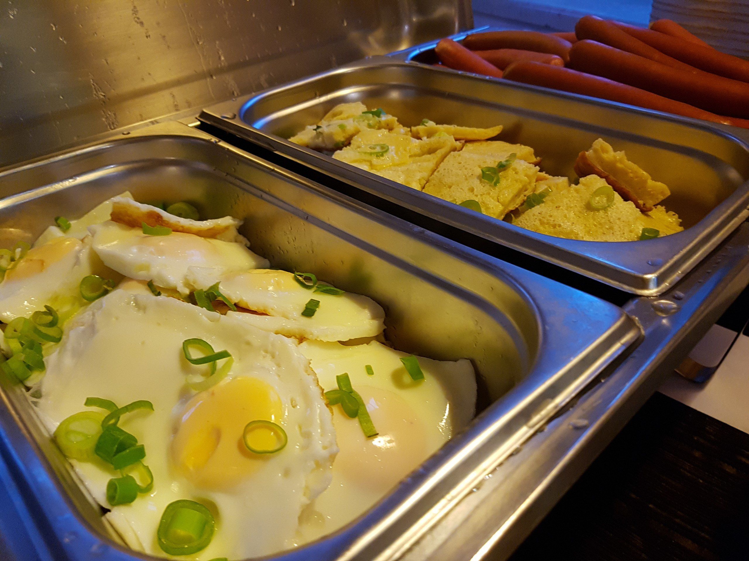 Buffet com diversos alimentos, incluindo ovos, cachorros-quentes e outros pratos.