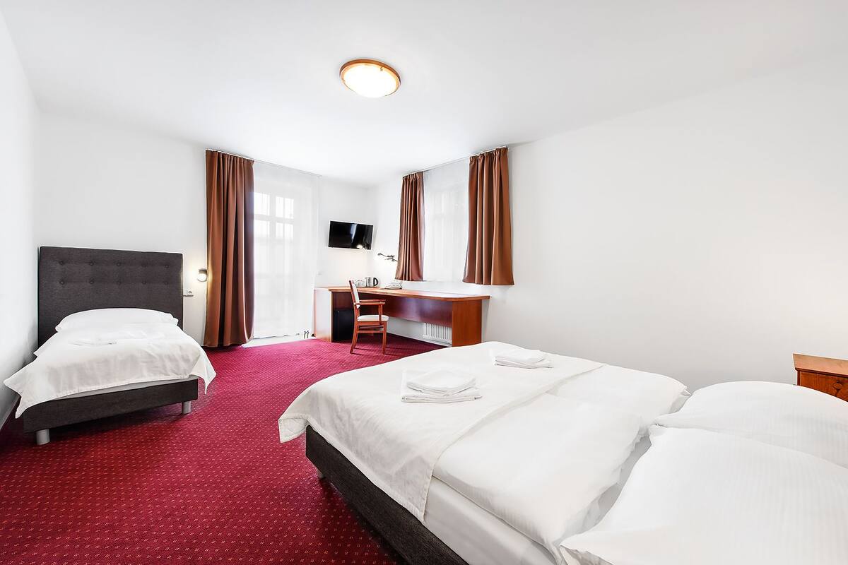 Quarto de hotel com duas camas de solteiro, uma escrivaninha, uma cadeira, uma televisão e um tapete vermelho.