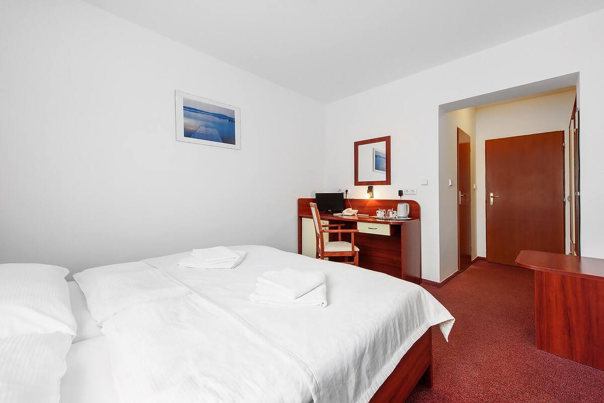 Quarto de hotel com uma cama, uma escrivaninha com cadeira, uma mesinha, um quadro na parede e uma porta.