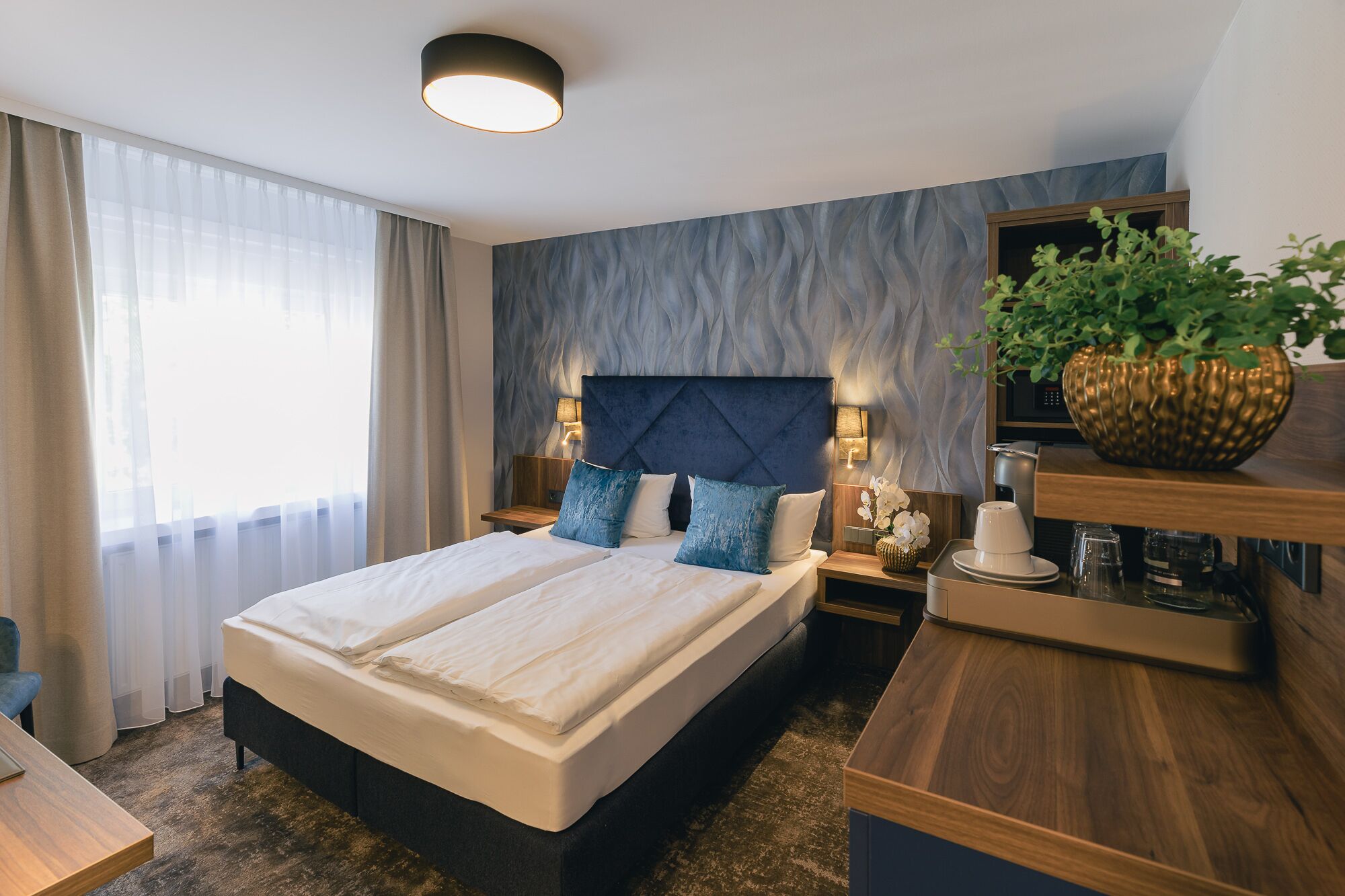 Quarto de hotel moderno com uma cama grande, mesas de cabeceira, um espelho e um vaso de planta.