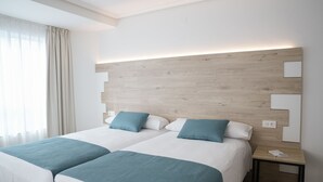 Quarto de hotel com duas camas, cabeceira de madeira, mesa de cabeceira e janela com cortinas.