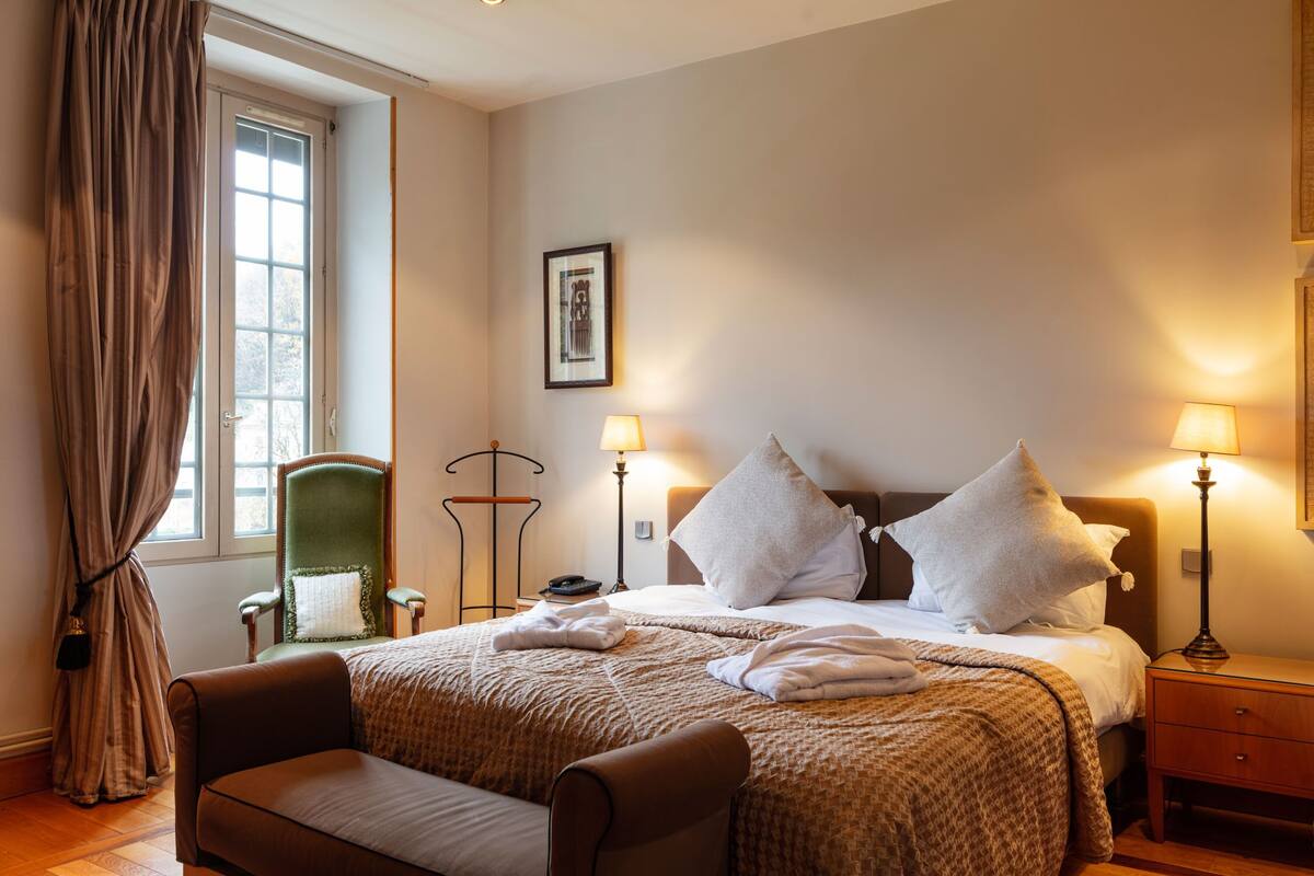 Quarto de hotel com uma cama grande, duas luminárias de cabeceira, uma cadeira, mesa de cabeceira e uma janela com cortinas.