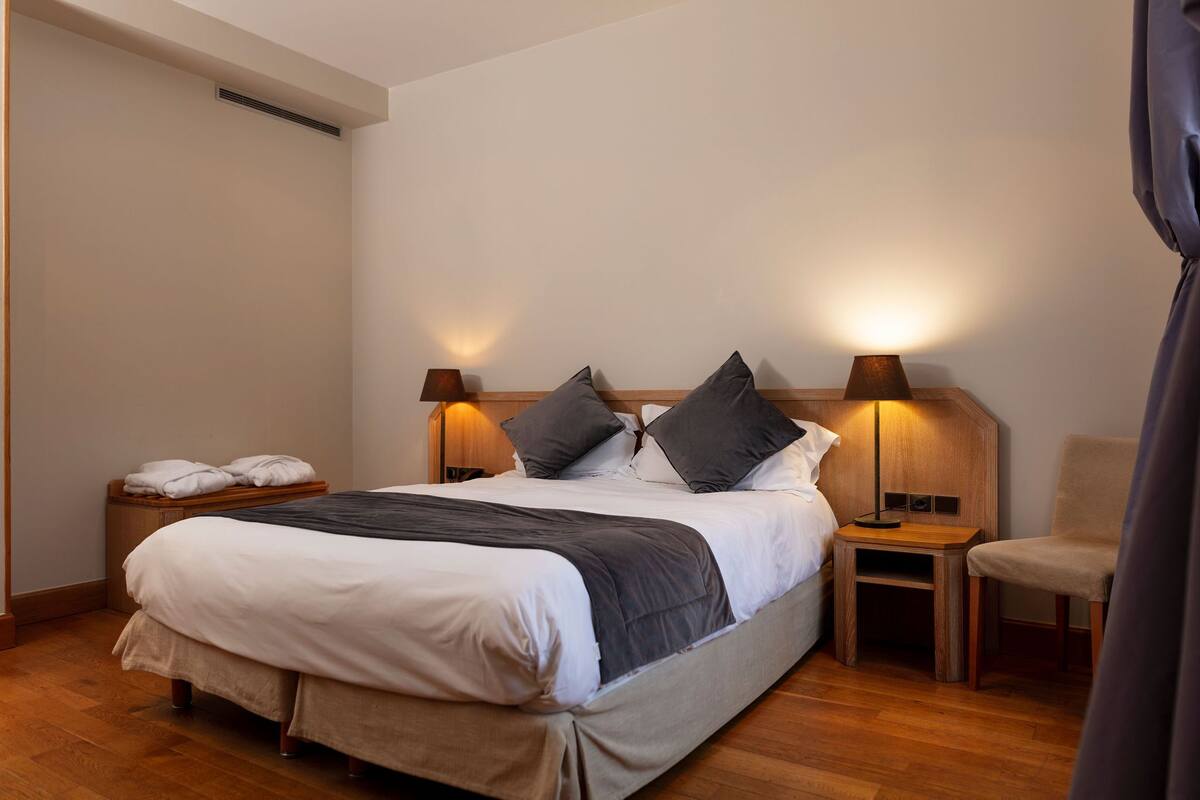 Uma cama bem arrumada com almofadas, uma mesa de cabeceira com abajur e uma cadeira em um quarto de hotel.