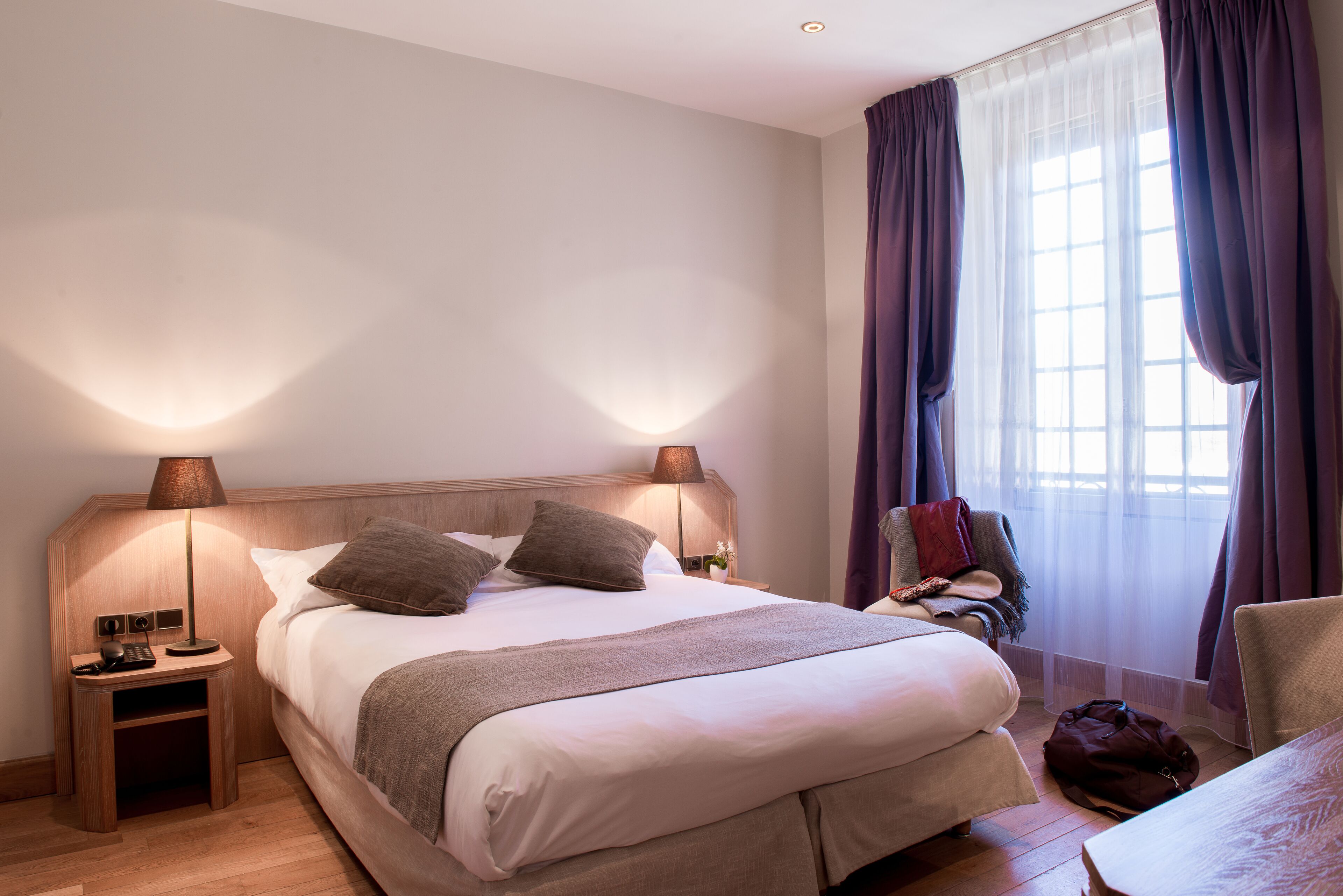 Quarto de hotel bem iluminado com uma cama grande, duas luminárias de cabeceira, mesa de cabeceira, uma cadeira e uma janela com cortinas.