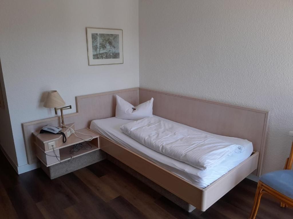 Uma cama de solteiro com estrutura de madeira, uma mesa de cabeceira com abajur e uma cadeira.