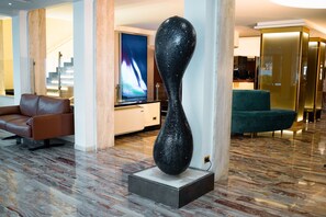 Um lobby moderno com uma grande escultura abstrata, um sofá de couro, uma poltrona verde e uma escada ao fundo.
