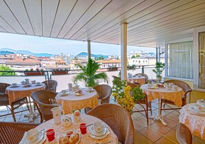 Terraço com mesas redondas e cadeiras de vime, com vista para um horizonte urbano que inclui edifícios e montanhas.