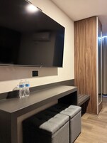 Uma sala de estar moderna com uma TV de tela plana na parede, um móvel de entretenimento cinza com duas garrafas de água e um painel de madeira à direita.