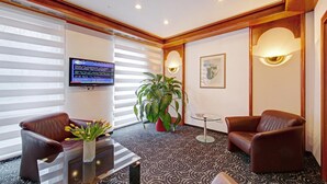 Um saguão de hotel com duas poltronas de couro, uma mesa de centro de vidro, um vaso de planta, uma pequena mesa redonda, uma televisão montada na parede e um quadro emoldurado na parede.