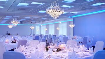 Salão de banquetes com várias mesas montadas para um evento formal, com lustres e toalhas de mesa brancas.