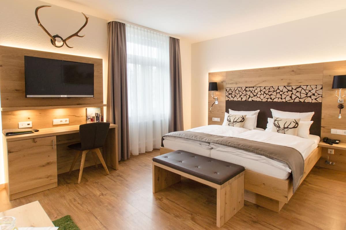 Quarto de hotel moderno com escrivaninha de madeira, cama confortável, um banco, televisão e uma grande janela com cortinas.