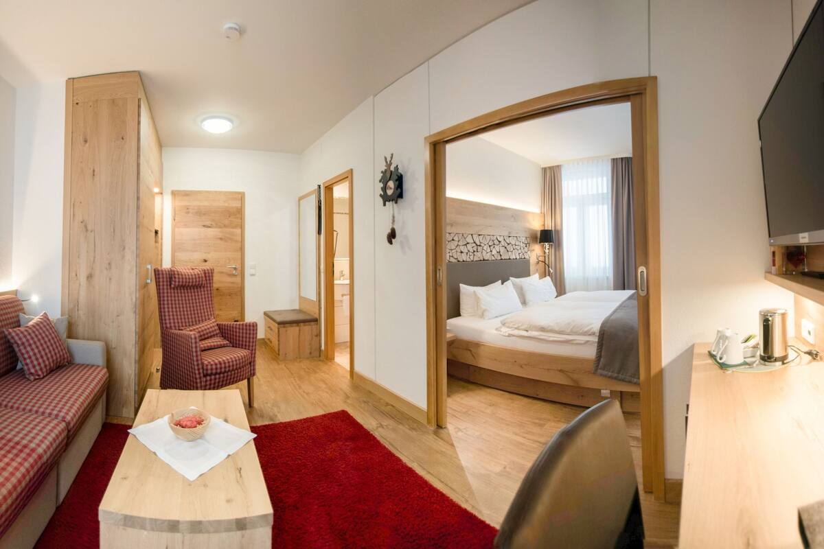 Quarto de hotel com sofá xadrez, mesa de centro de madeira, tapete vermelho, cama com roupa de cama branca, espelho e pintura de montanhas na parede.