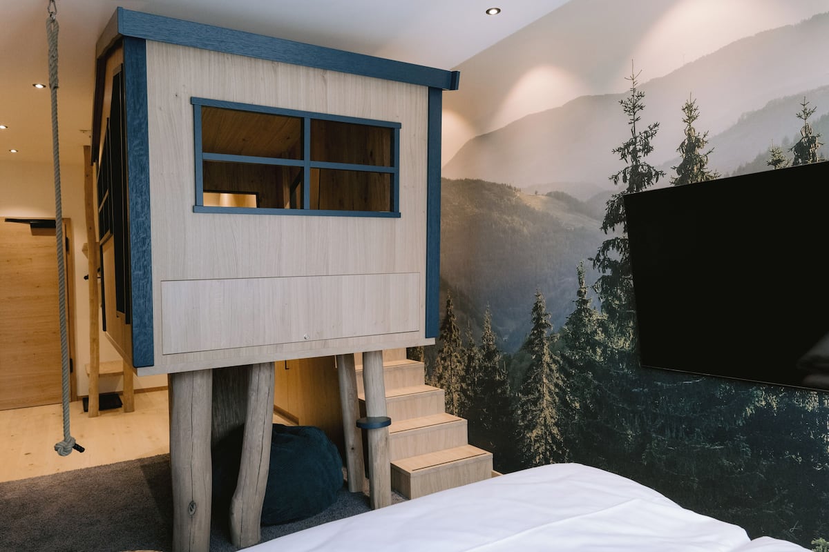 Quarto com cama estilo loft, casinha de madeira e um mural de floresta.