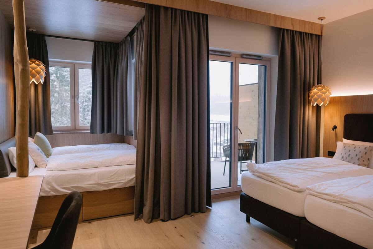 Quarto de hotel com duas camas, uma escrivaninha de madeira, uma cadeira, uma varanda com mesa e cortinas.