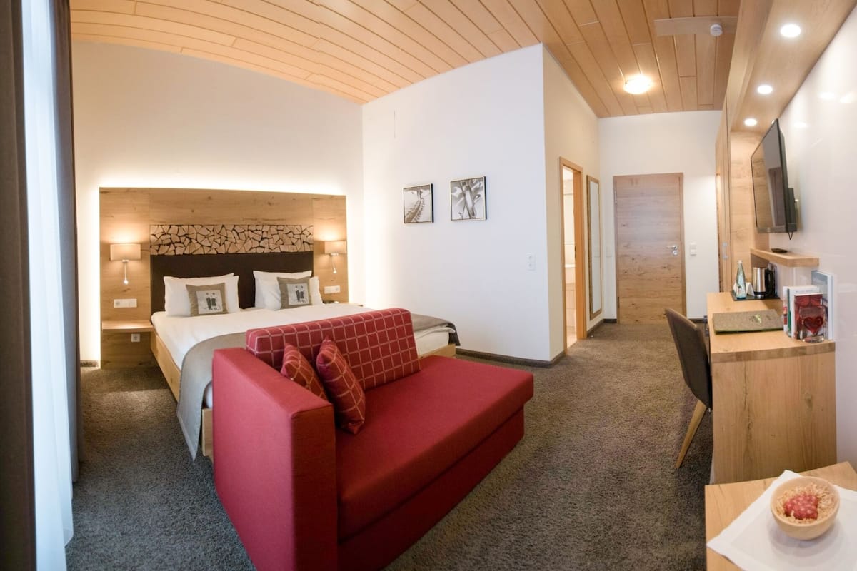 Quarto de hotel moderno com teto de madeira, cama com cabeceira, sofá vermelho, escrivaninha com cadeira e televisão.