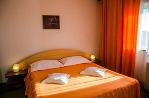Quarto de hotel com cama, duas luminárias de cabeceira, quadro na parede e cortinas laranja.
