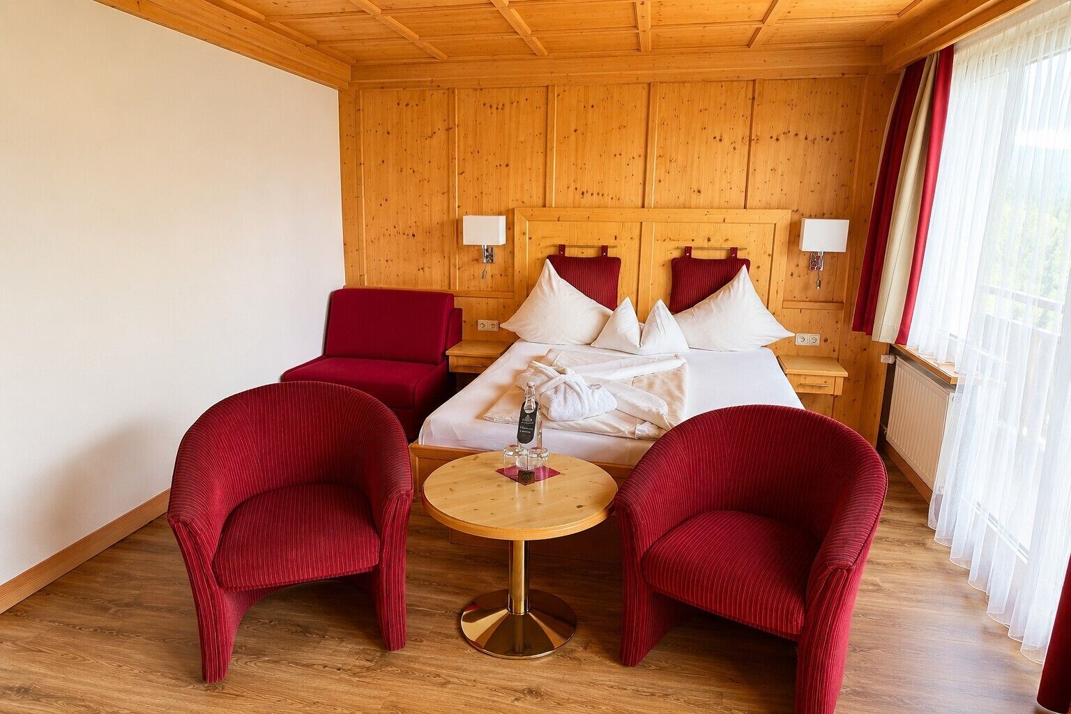 Quarto de hotel com paredes em painel de madeira, uma cama com lençóis brancos, duas poltronas vermelhas, uma mesa redonda pequena com uma garrafa e copos, e uma janela com cortinas vermelhas.