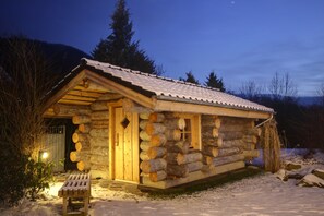 Uma cabana de tronco com porta e janela de madeira, rodeada por neve e árvores.