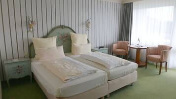 Quarto de hotel com uma cama de casal, duas mesas de cabeceira, uma mesinha redonda com uma cadeira e uma janela grande com cortinas.