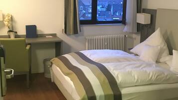 Quarto de hotel com cama, escrivaninha, radiador e janela com cortinas.