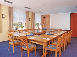 Uma sala de conferência com uma mesa comprida, várias cadeiras, uma tela de projetor e uma janela grande com cortinas.