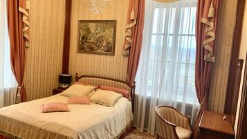 Quarto com cama grande, lustre, cortinas, pintura, cadeira e escrivaninha.