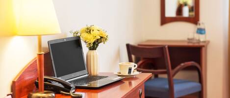 Mesa de quarto de hotel com um laptop, um vaso de flores, uma xícara de café, uma luminária e uma cadeira.