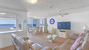 Sala de estar com uma televisão grande, sofás de couro, uma mesa de centro de vidro e vista para o oceano.