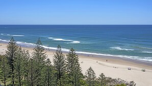 Uma praia com águas azul-claras, areal arenoso e altas árvores de pinheiro em primeiro plano.