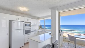 Cozinha moderna com armários brancos, geladeira, forno e uma ilha grande. Há uma varanda com vista para a praia e o oceano.
