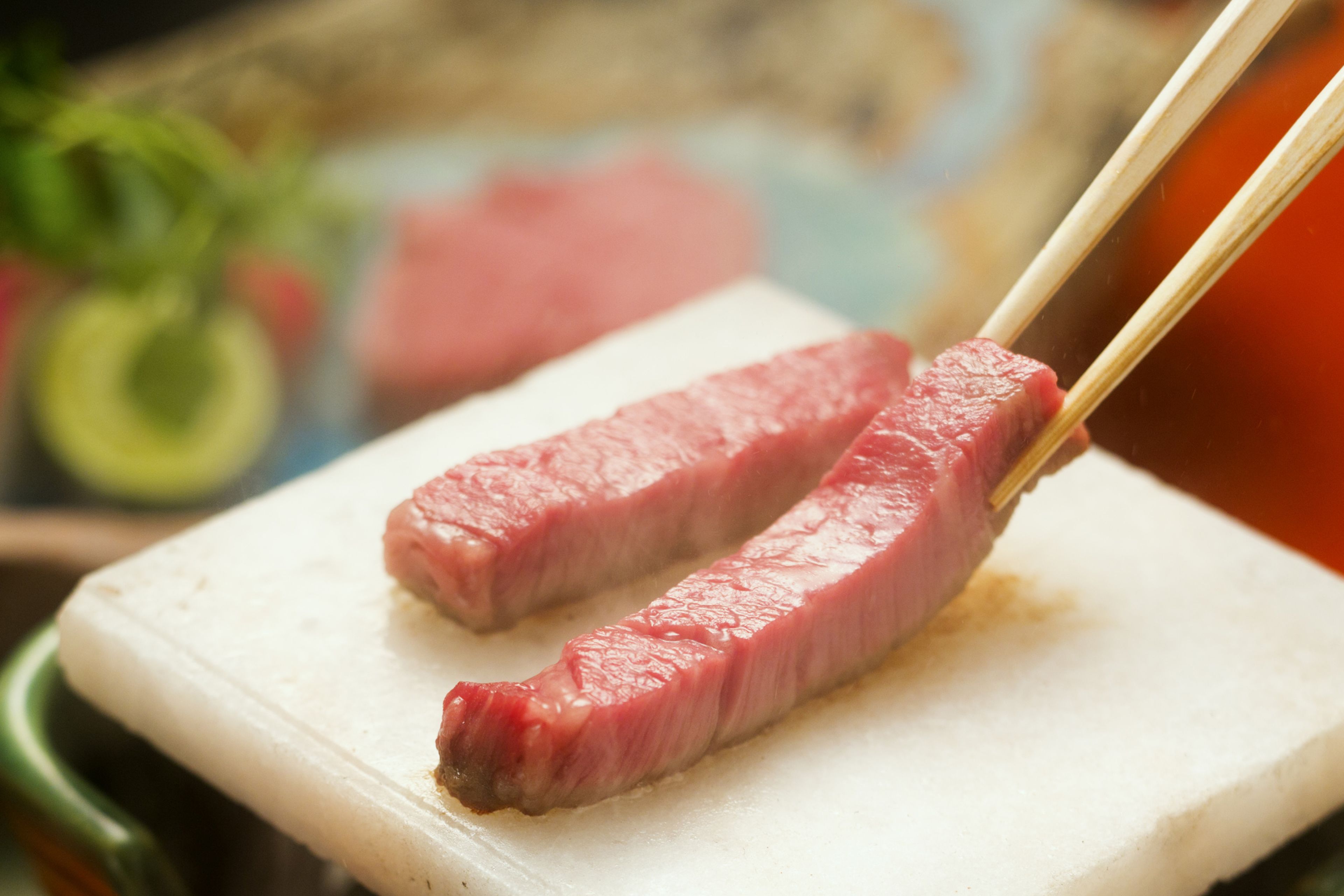 Close-up de um pedaço de carne crua em tábua de corte branca, com pauzinhos sobrepostos.