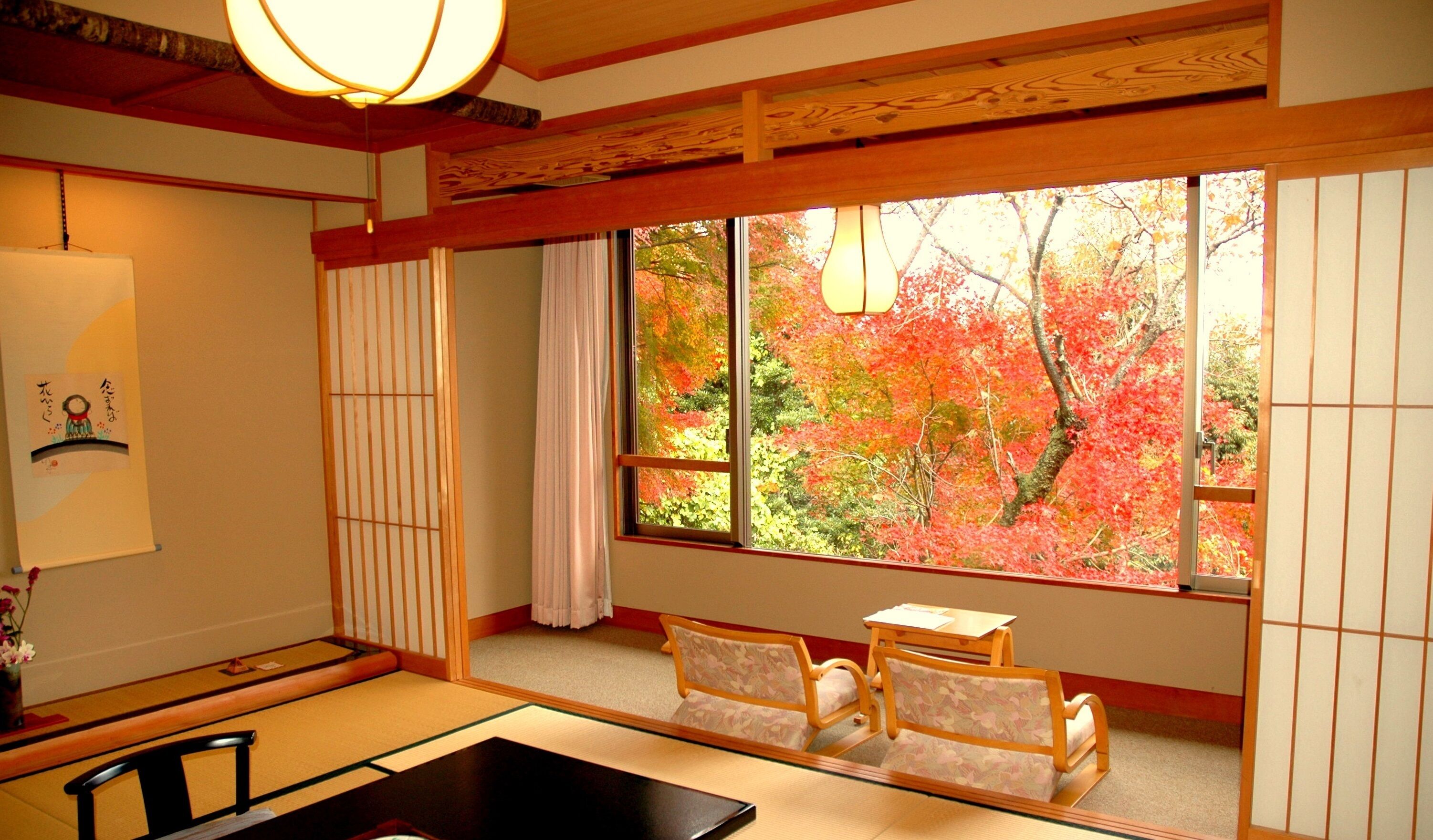 Um quarto japonês tradicional com tatames, uma mesa baixa, duas cadeiras, uma janela com vista para folhagem outonal e um lustre.