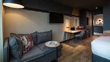 Quarto de hotel moderno com um sofá cinzento, uma mesinha redonda com uma revista, uma escrivaninha com um computador, uma cama e um espelho grande com luzes circulares.