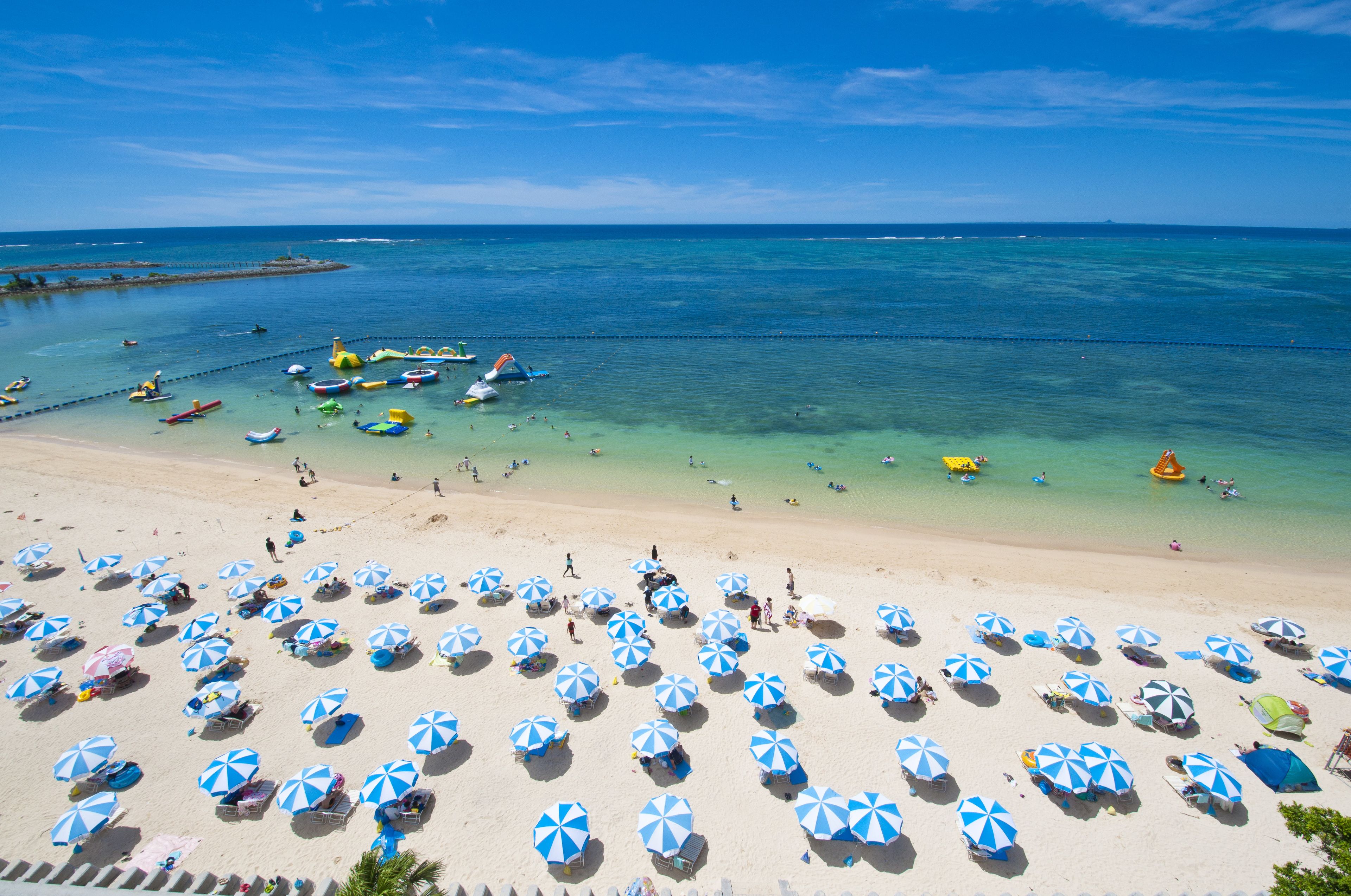 Uma praia com vários guarda-sóis azuis e brancos, água turquesa transparente e uma costa arenosa.