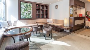 Um hall de hotel moderno com sofá, mesa redonda e cadeiras, uma janela ampla com arte de plantas e um balcão de recepção.