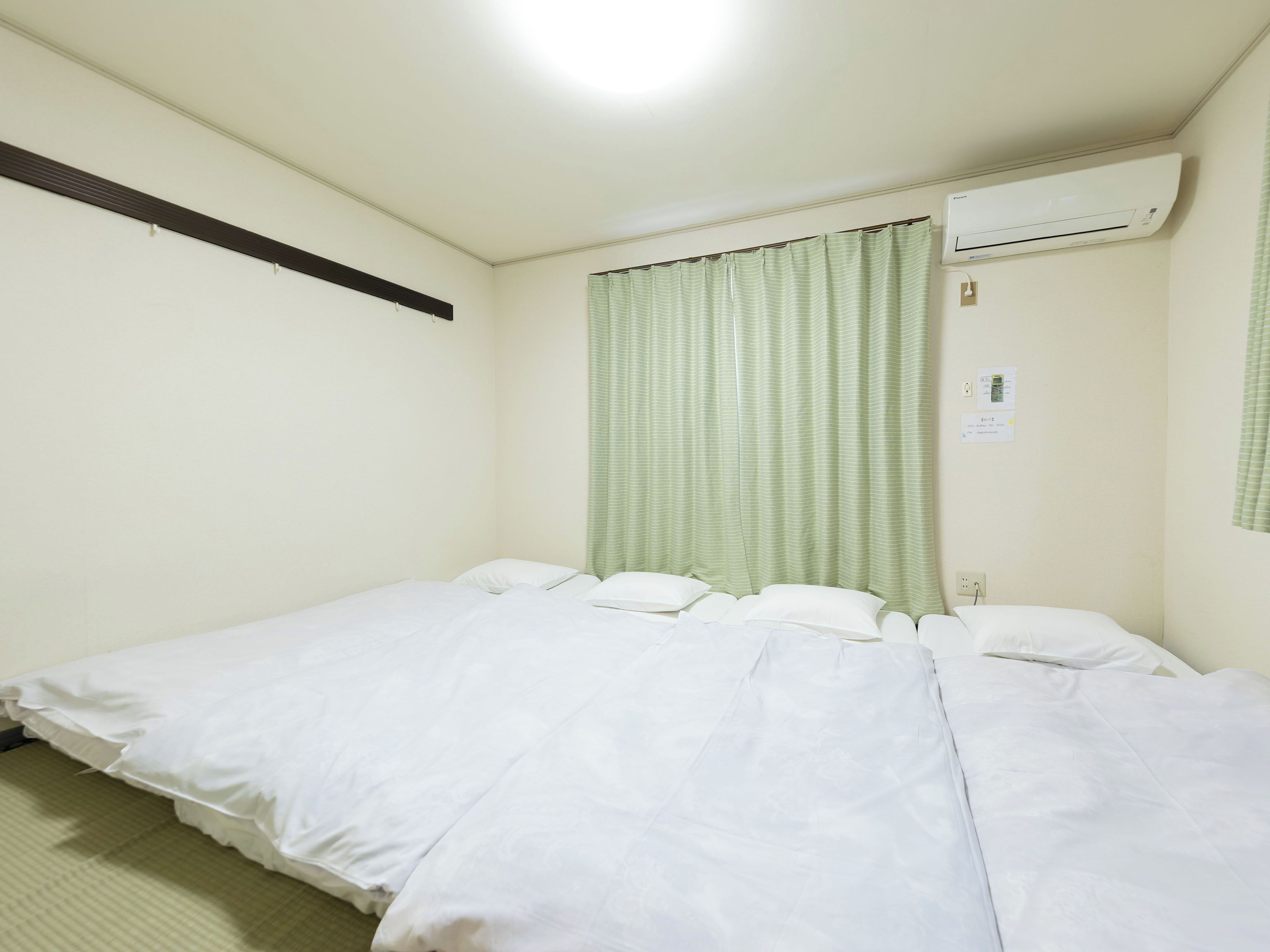 Uma cama de solteiro com roupa de cama branca em um quarto com cortina verde e um aparelho de ar condicionado na parede.