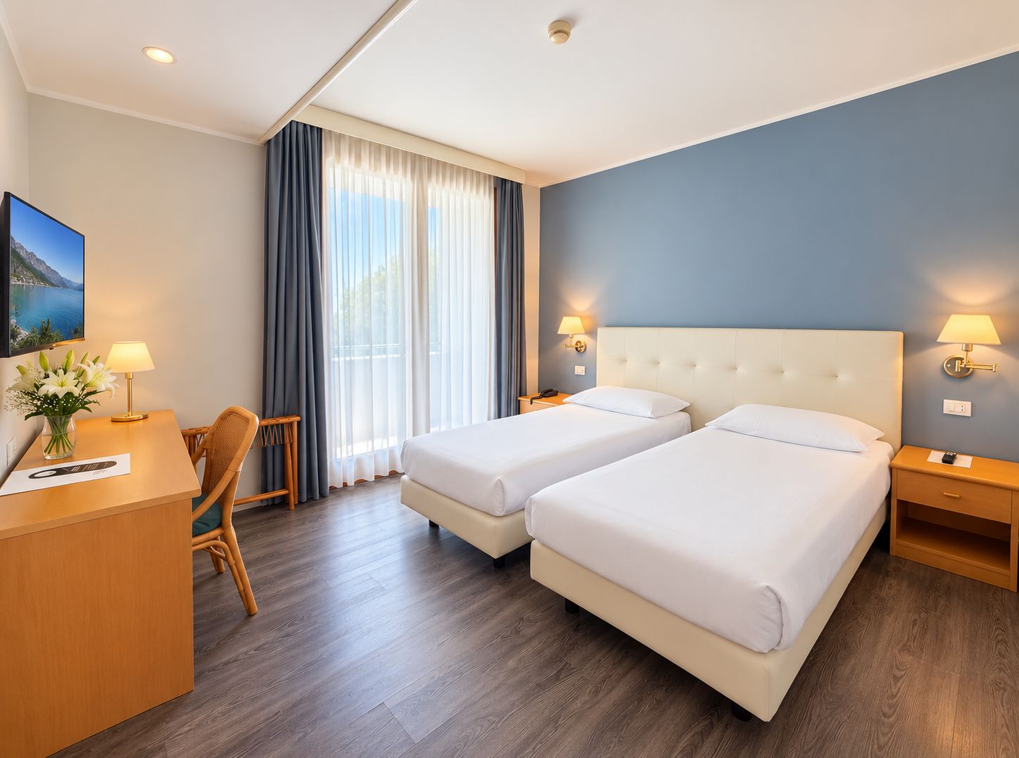 Quarto de hotel com duas camas, uma escrivaninha, uma cadeira e uma janela grande com cortinas.