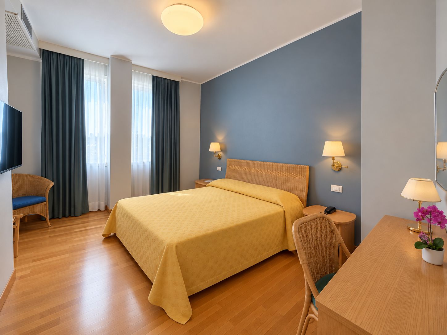 Quarto de hotel com cama, escrivaninha, cadeira, televisão e planta em vaso.