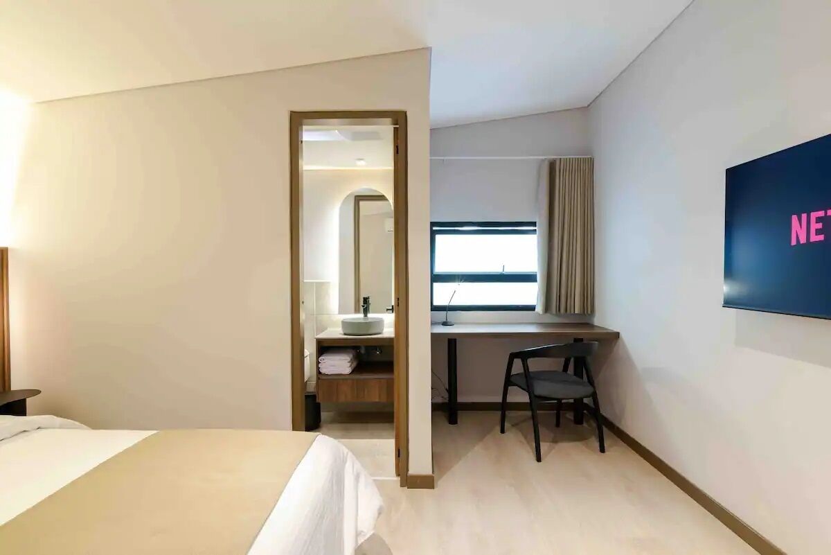 Quarto de hotel com cama, escrivaninha, cadeira, televisão e banheiro visível por uma porta aberta.